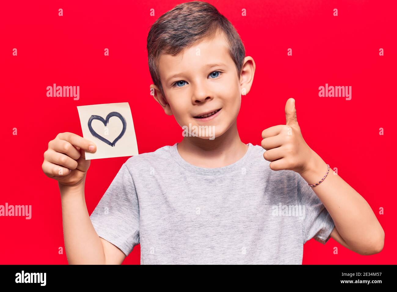 Cute blond kid holding heart reminder smiling happy and positive, thumb ...