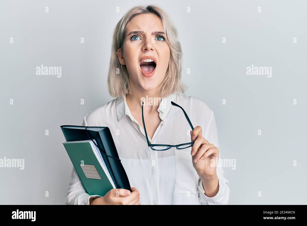Young blonde girl reading a book and holding glasses angry and mad ...