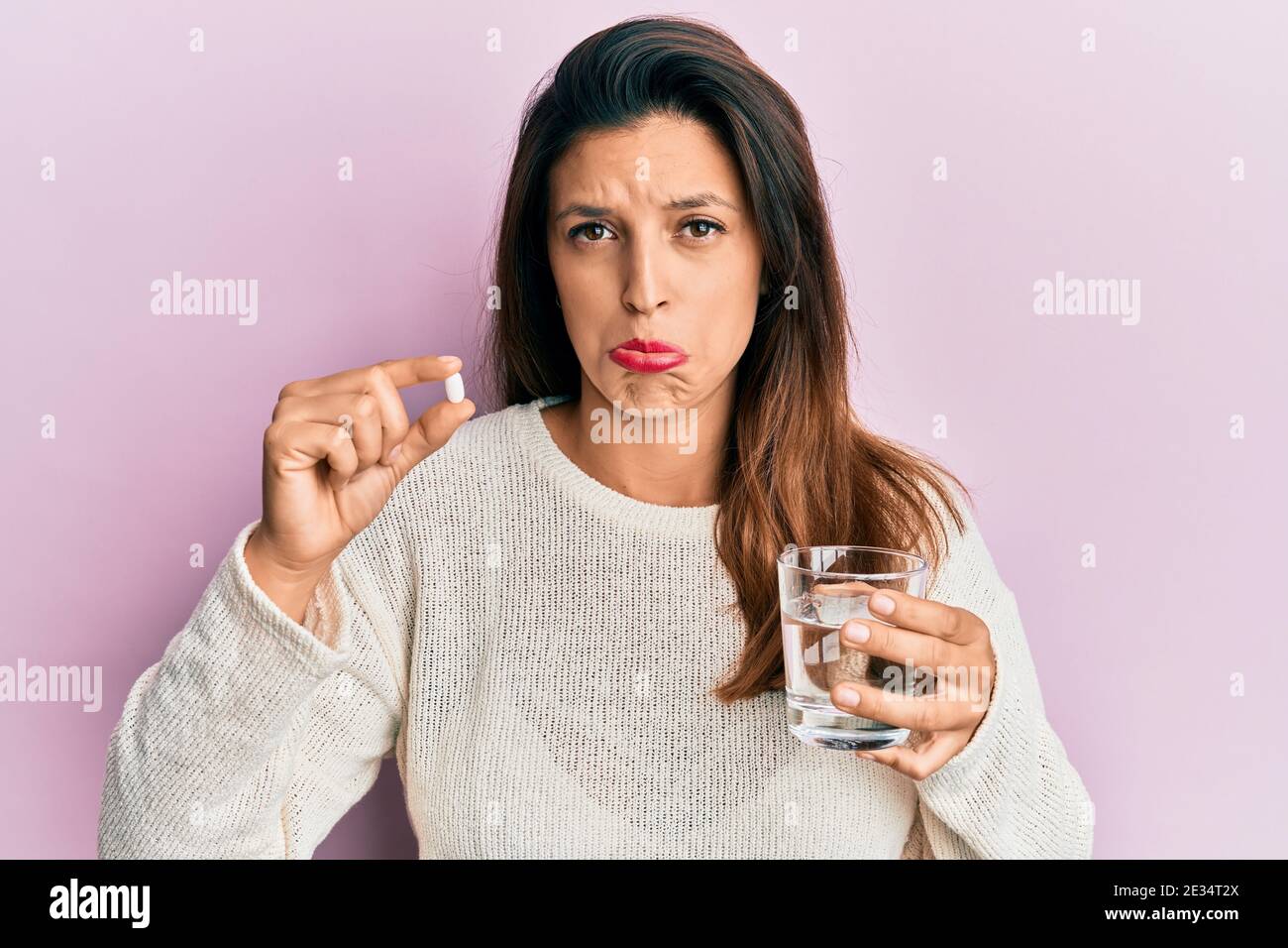 Beautiful hispanic woman holding pill and glass of water depressed and ...