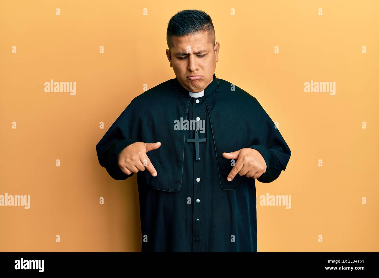 Young latin priest man standing over yellow background pointing down ...