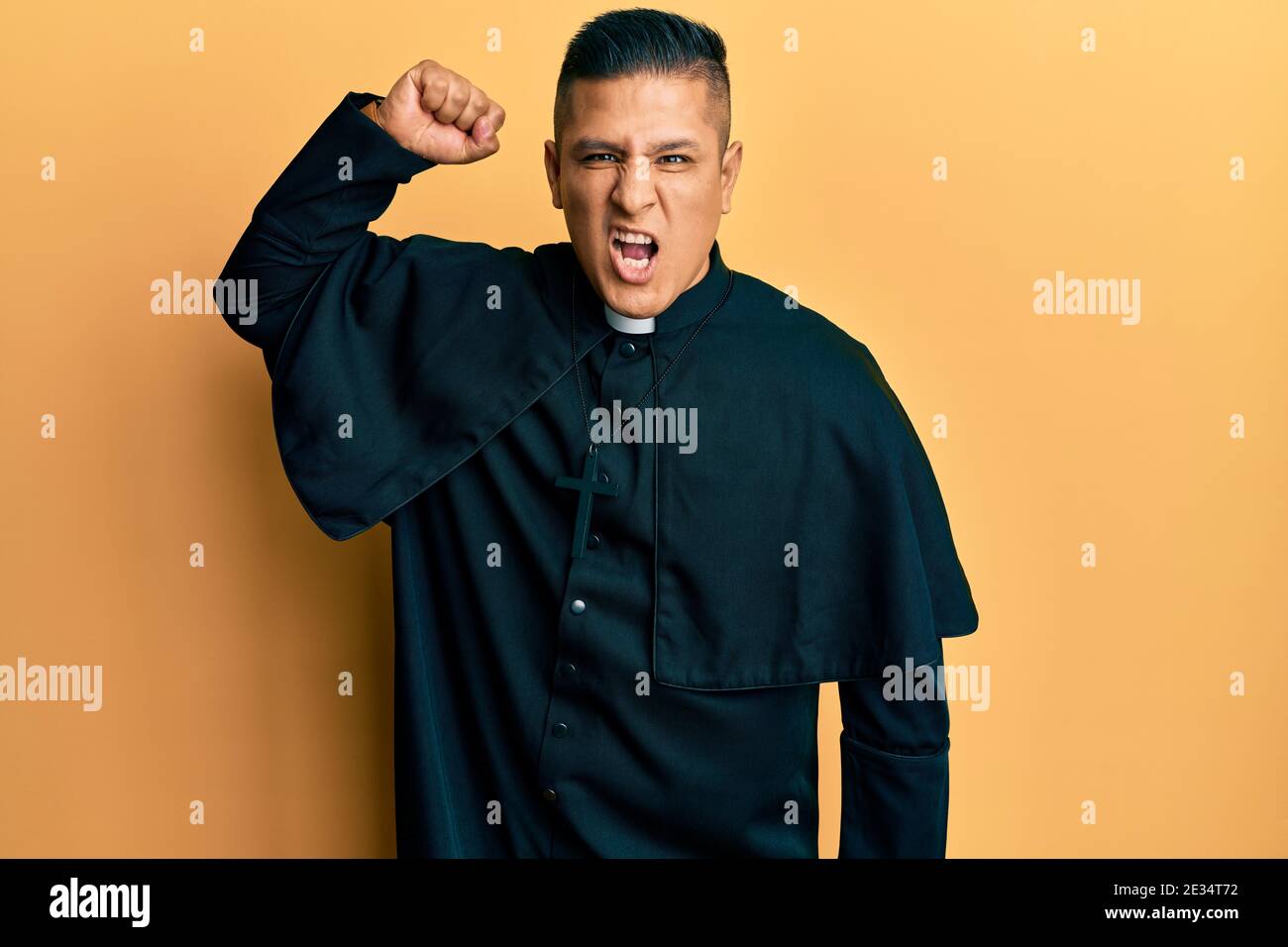 Young latin priest man standing over yellow background angry and mad ...