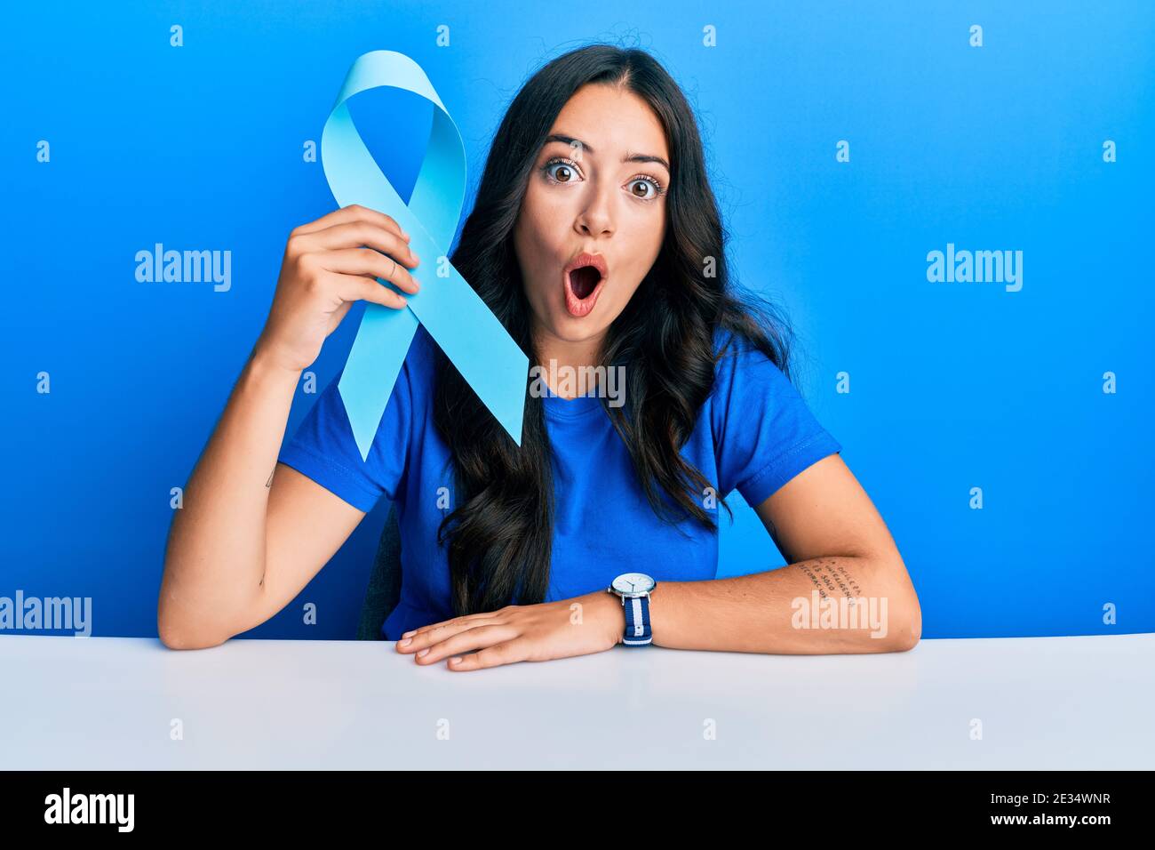 Beautiful brunette young woman holding blue ribbon scared and amazed ...