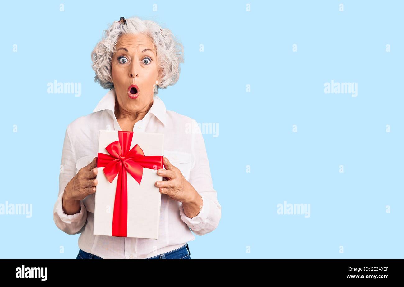 Senior grey-haired woman holding gift scared and amazed with open mouth ...