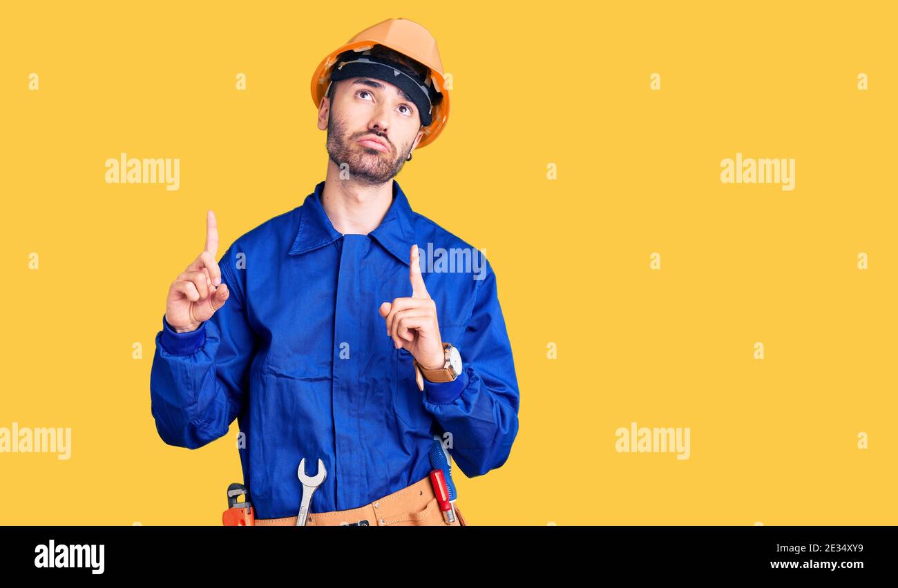 Young hispanic man wearing worker uniform pointing up looking sad and ...