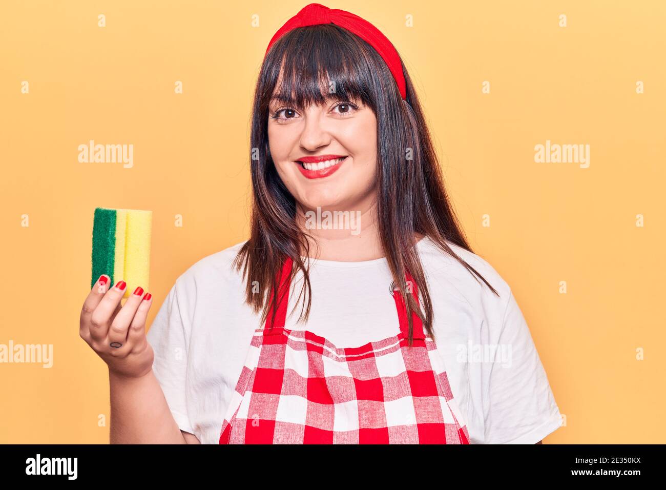 Young plus size woman wearing apron holding scourer looking positive ...