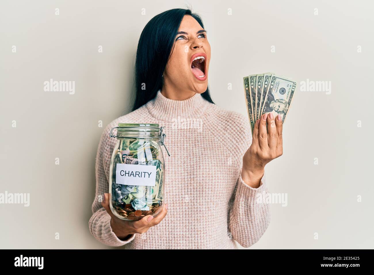 Young caucasian woman holding charity jar and dollars banknotes angry ...
