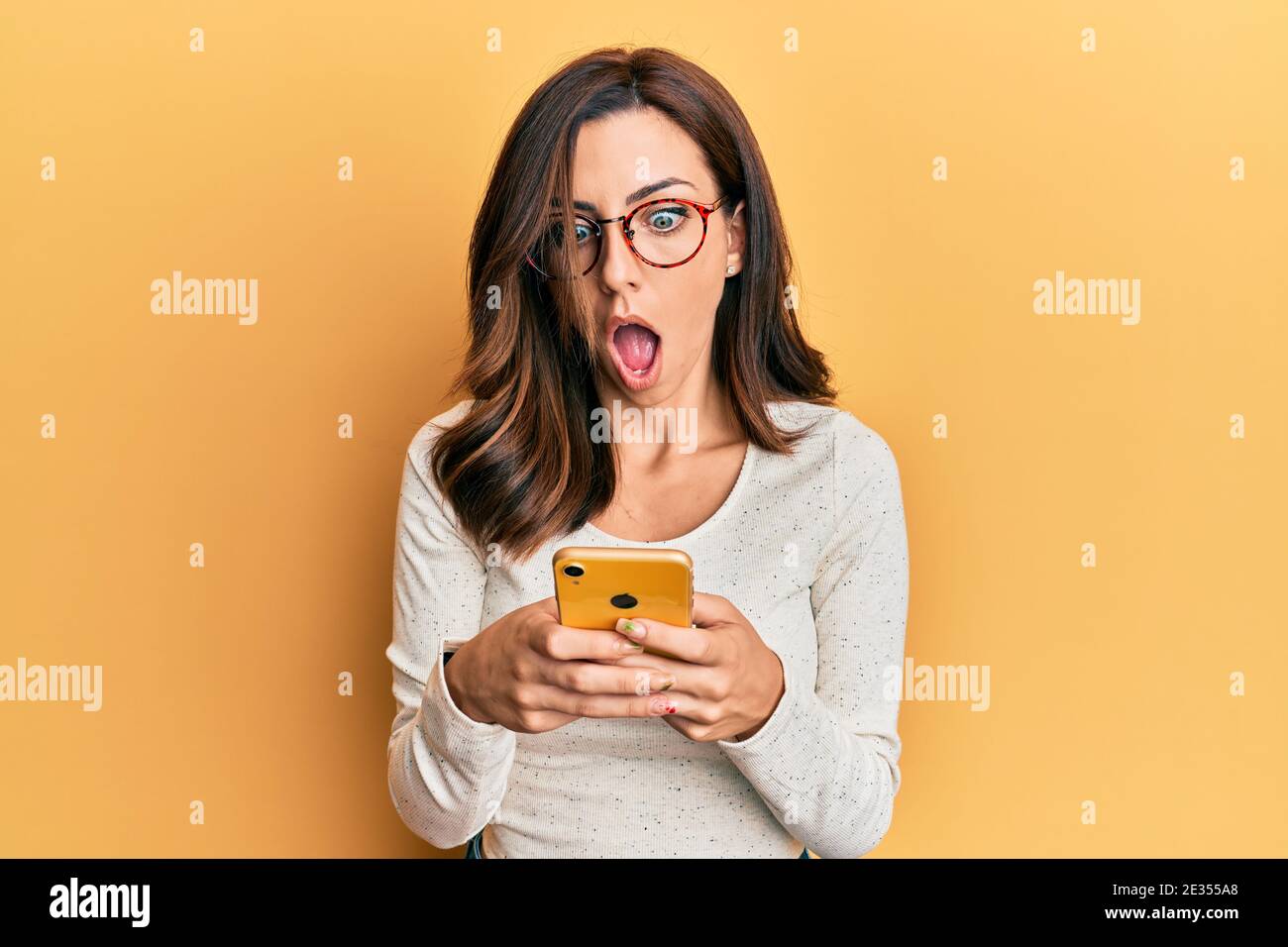 Young brunette woman using smartphone over yellow background afraid and ...