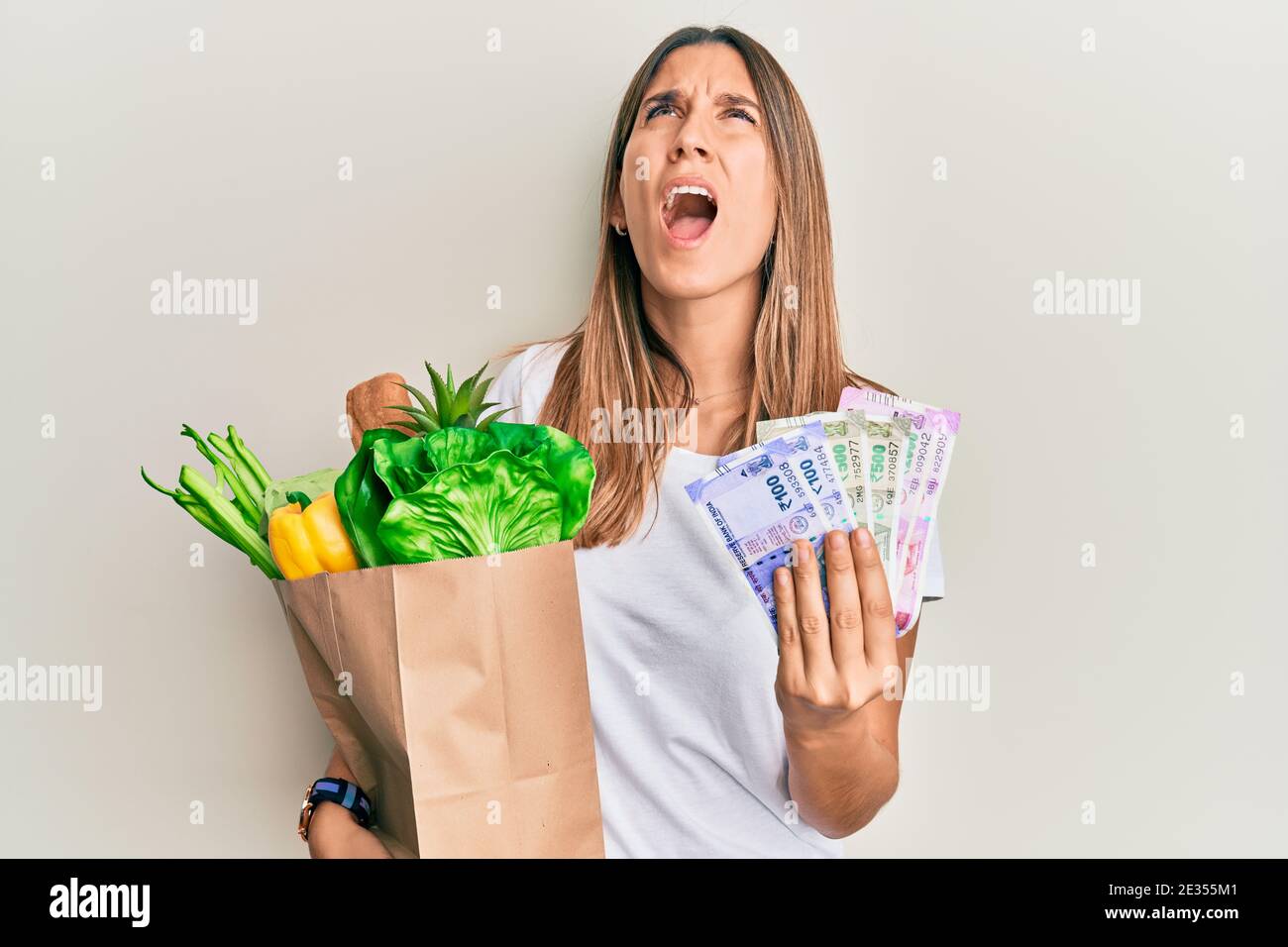 Brunette young woman holding groceries and indian rupee banknotes angry ...