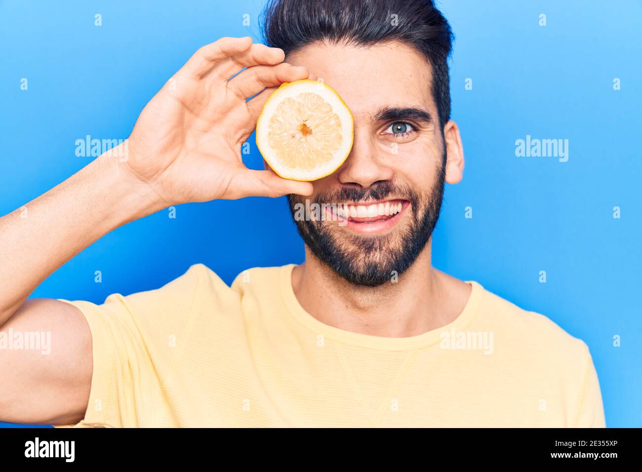 Young handsome man with beard holding slice of lemon over eye looking ...