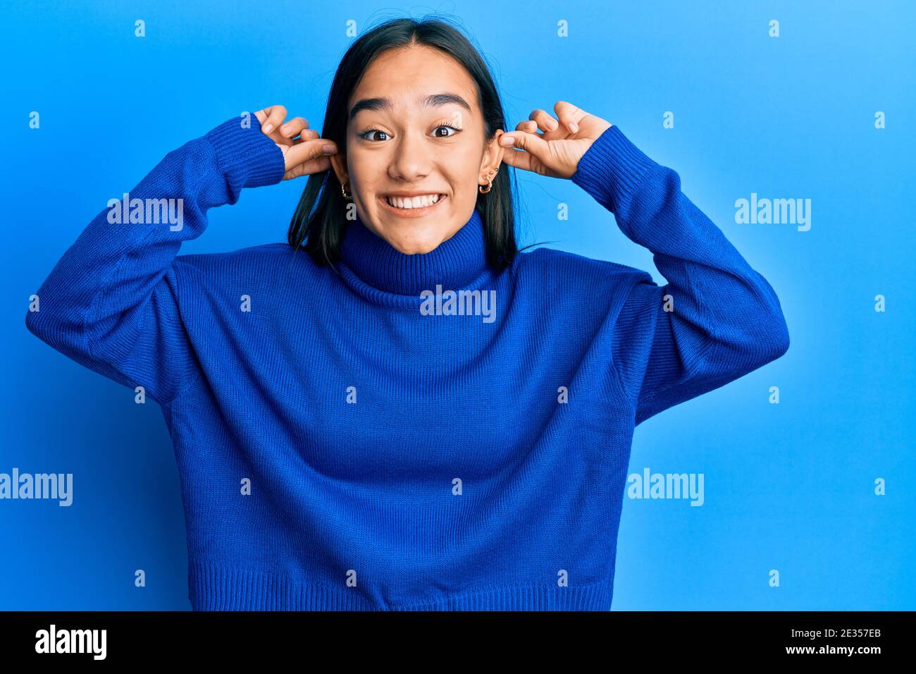 Young asian woman wearing casual winter sweater smiling pulling ears ...