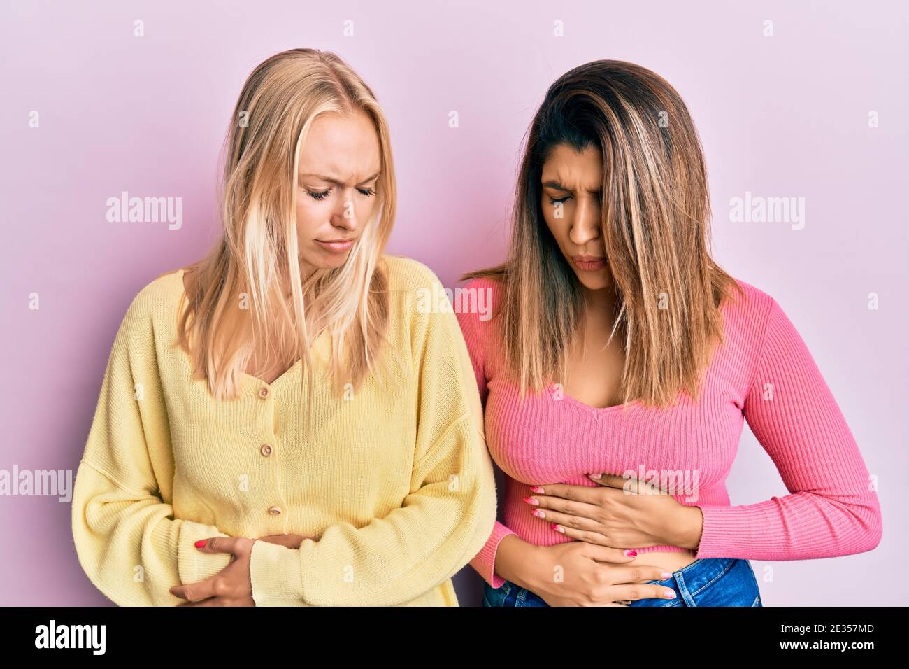 Two friends standing together over pink background with hand on stomach ...