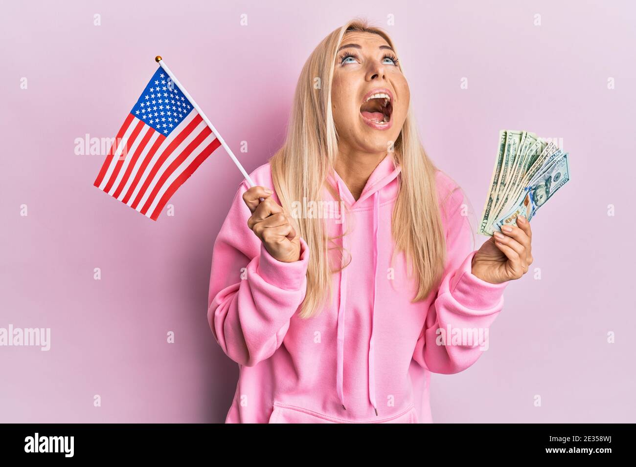 Young blonde woman holding united states flag and dollars angry and mad ...