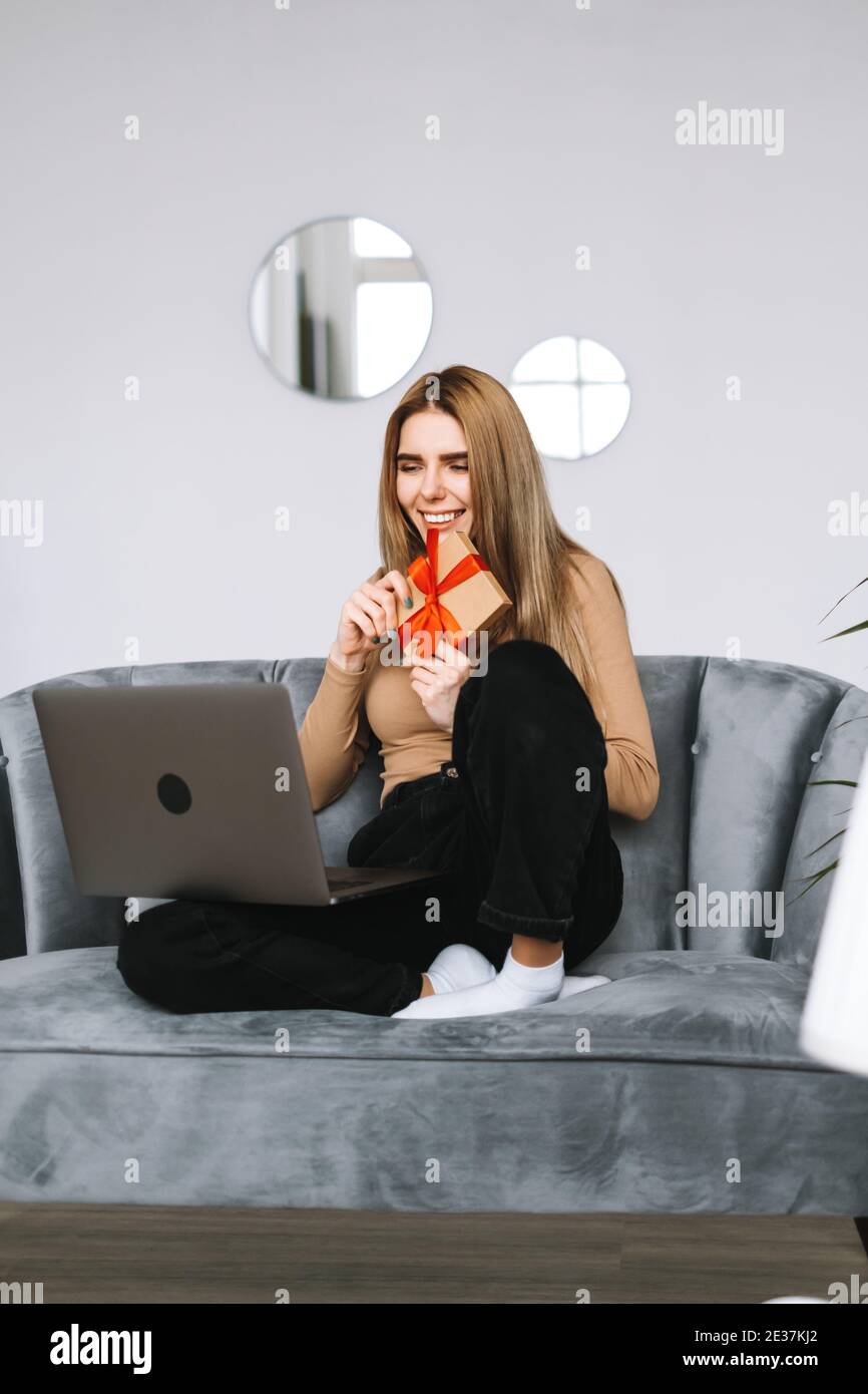 Happy young woman opening gift in front of laptop during video call or ...