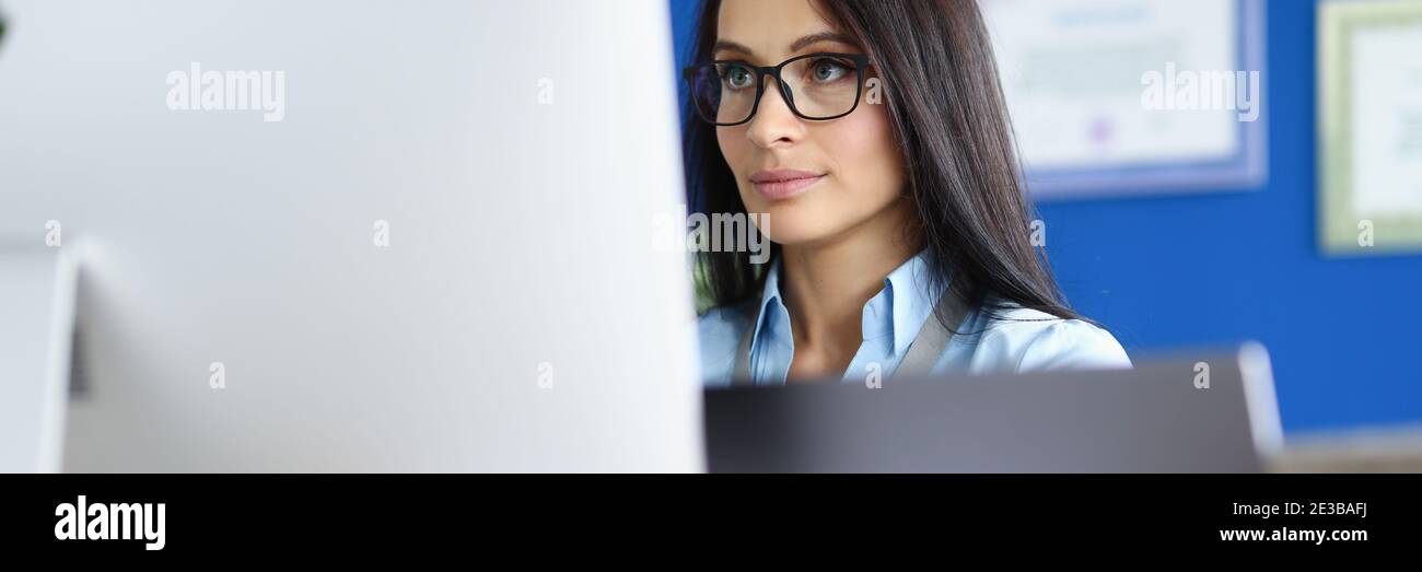 Woman in glasses and blue shirt sit at workplace and look at computer ...