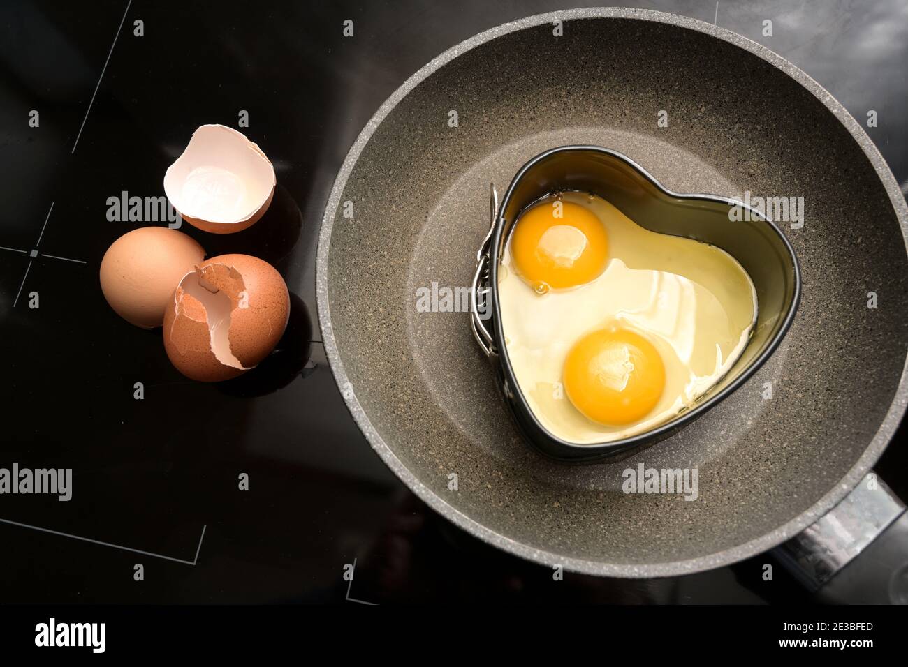 Two fried eggs in heart shape on a frying pan and empty shells on the black stove, cooking with ...