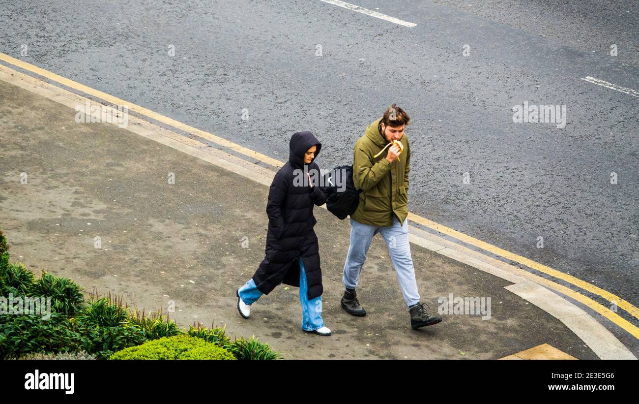 Couple walking down the road. Man eating banana and woman wearing long ...
