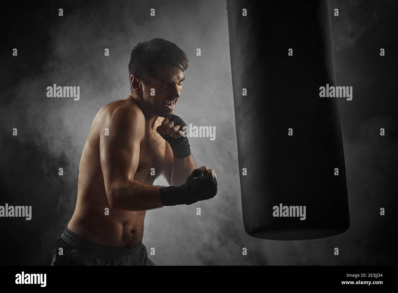 Aggressive boxer in black boxing wraps punching in boxing bag on dark ...