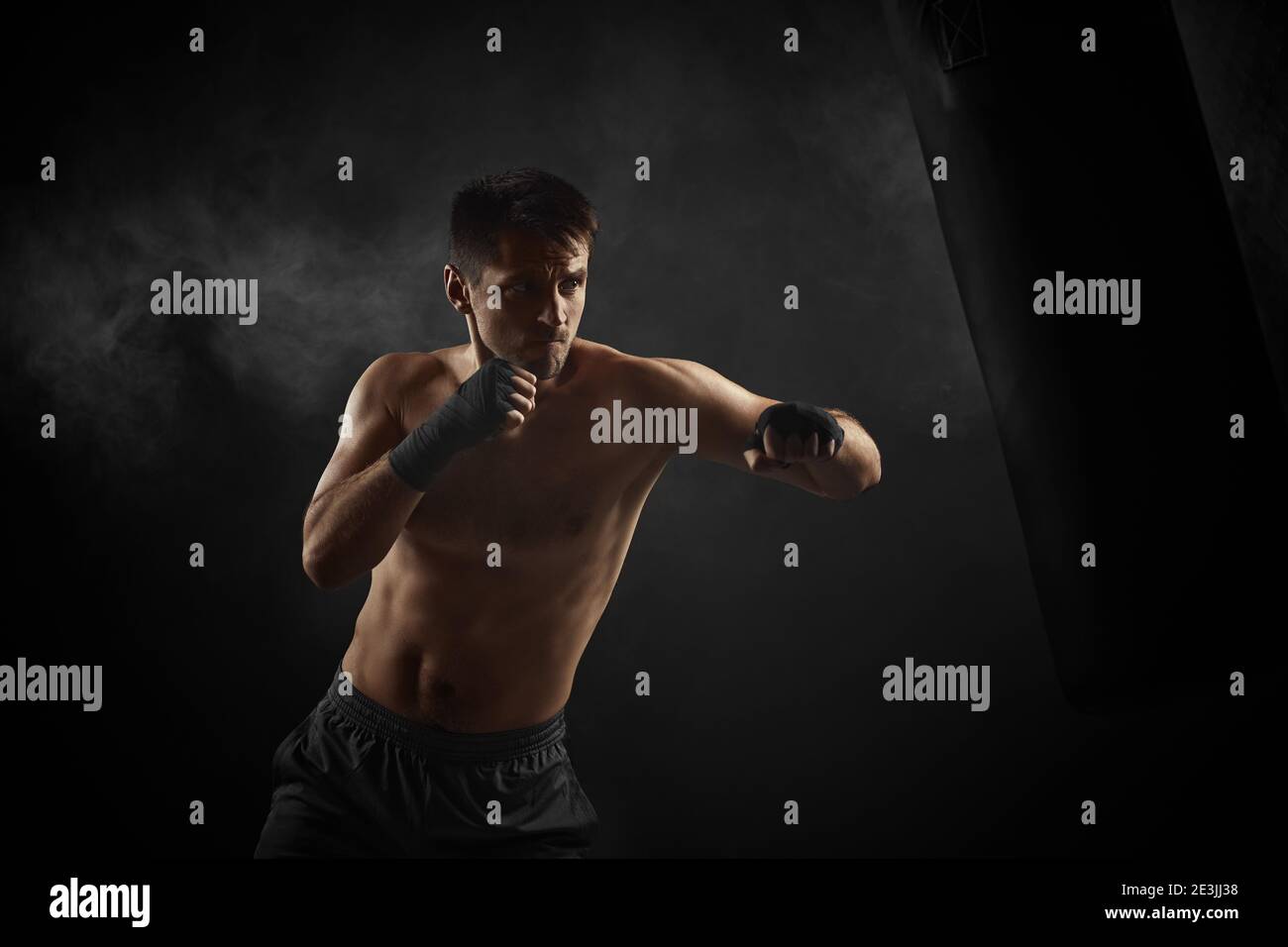 male boxer in black boxing wraps punching in boxing bag on dark ...