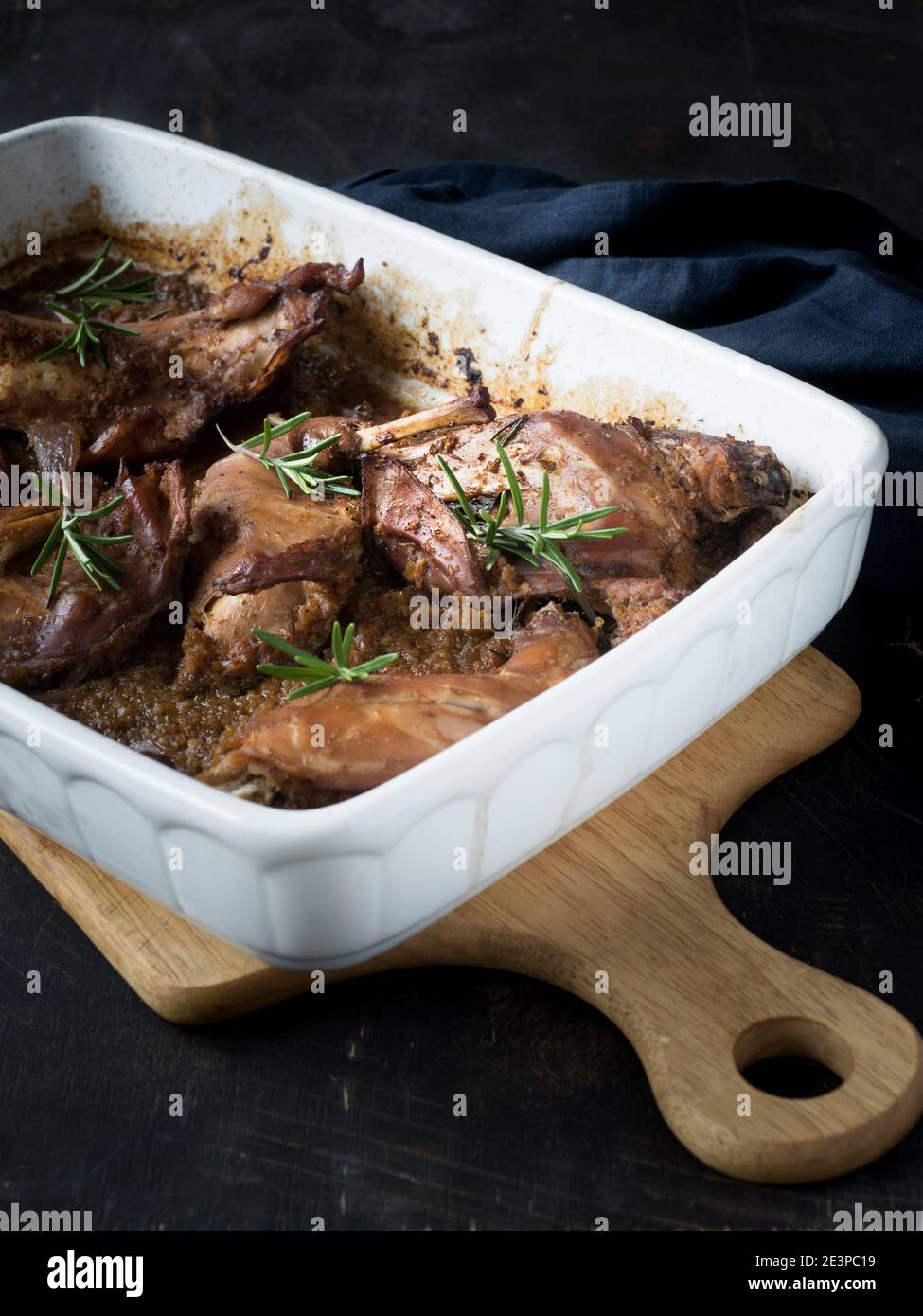 Roasted rabbit with rosemary in white pan on wooden cutting board. Dark ...