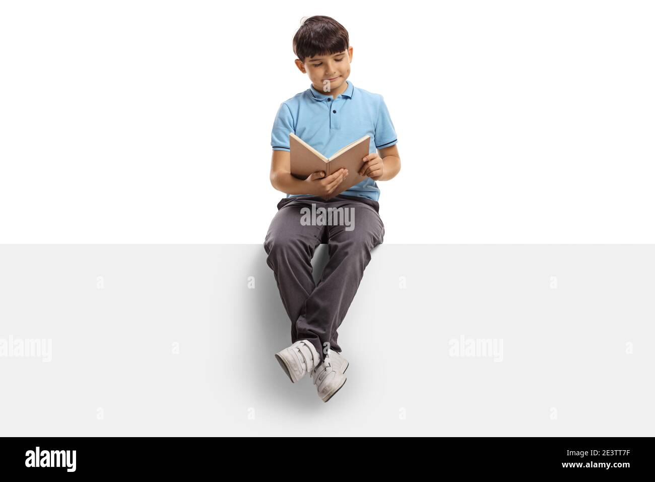 Boy sitting on a blank panel and reading a book isolated on white ...