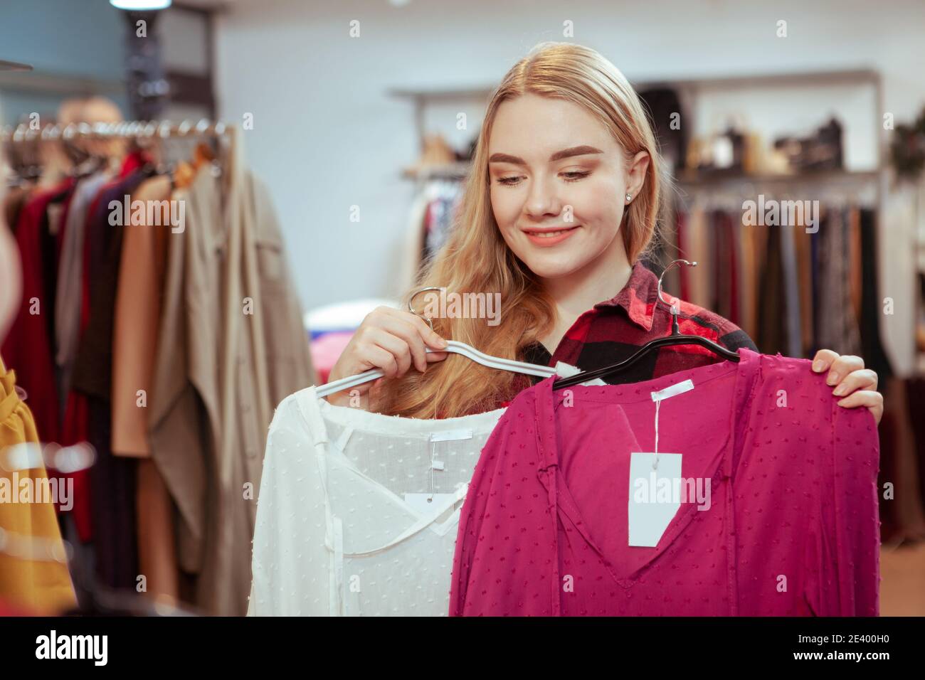 Attractive young woman smiling joyfully, choosing between two blouses ...