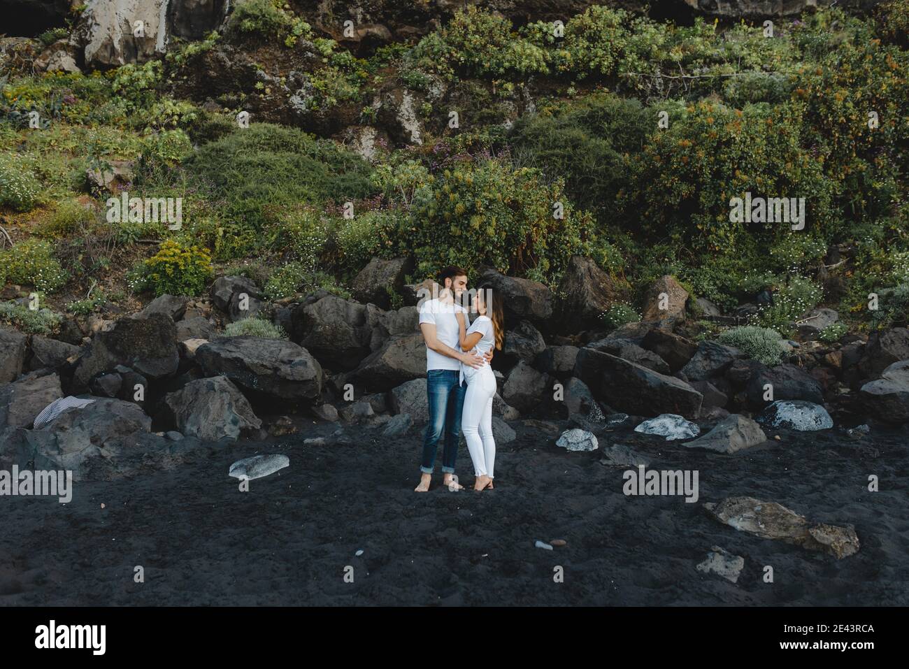 Side view of happy couple standing on sandy beach and cuddling while ...