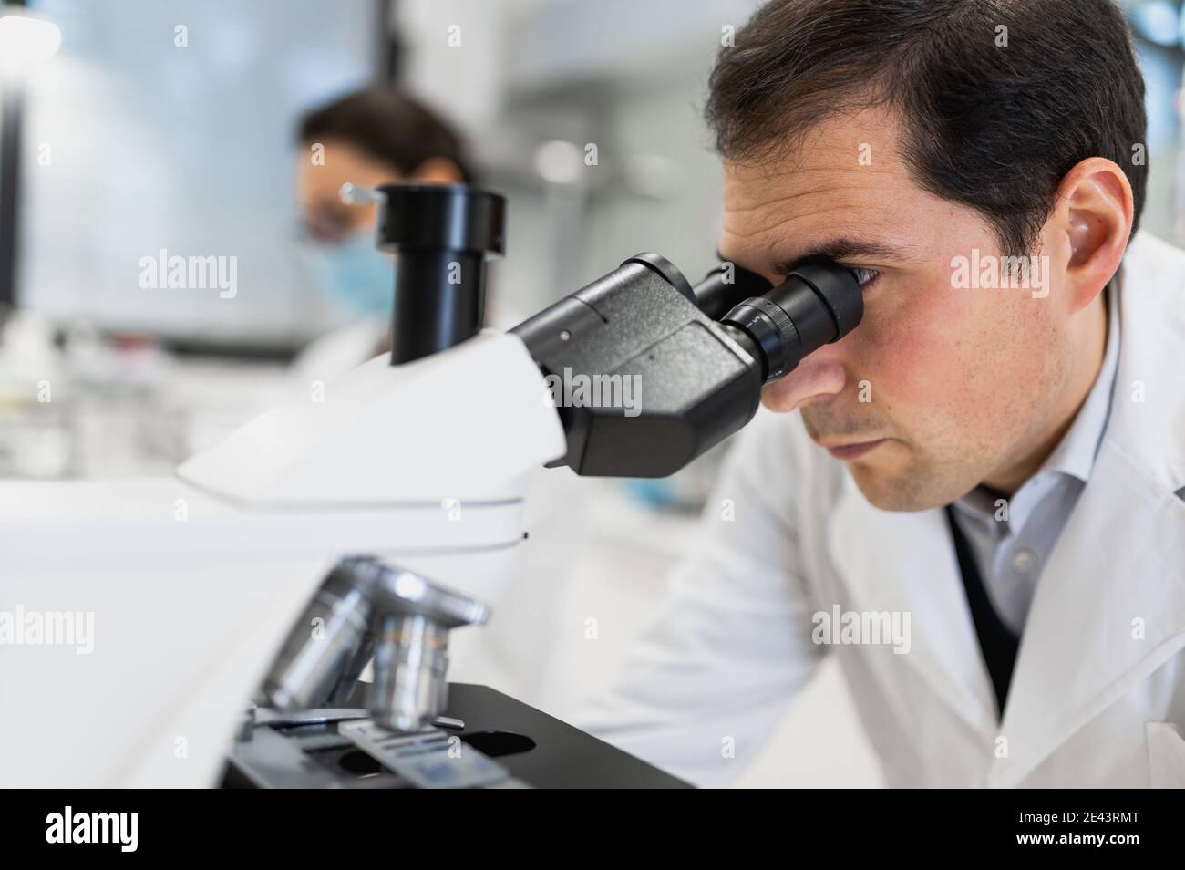 Concentrated male scientist using microscope and analyzing chemical samples while female ...