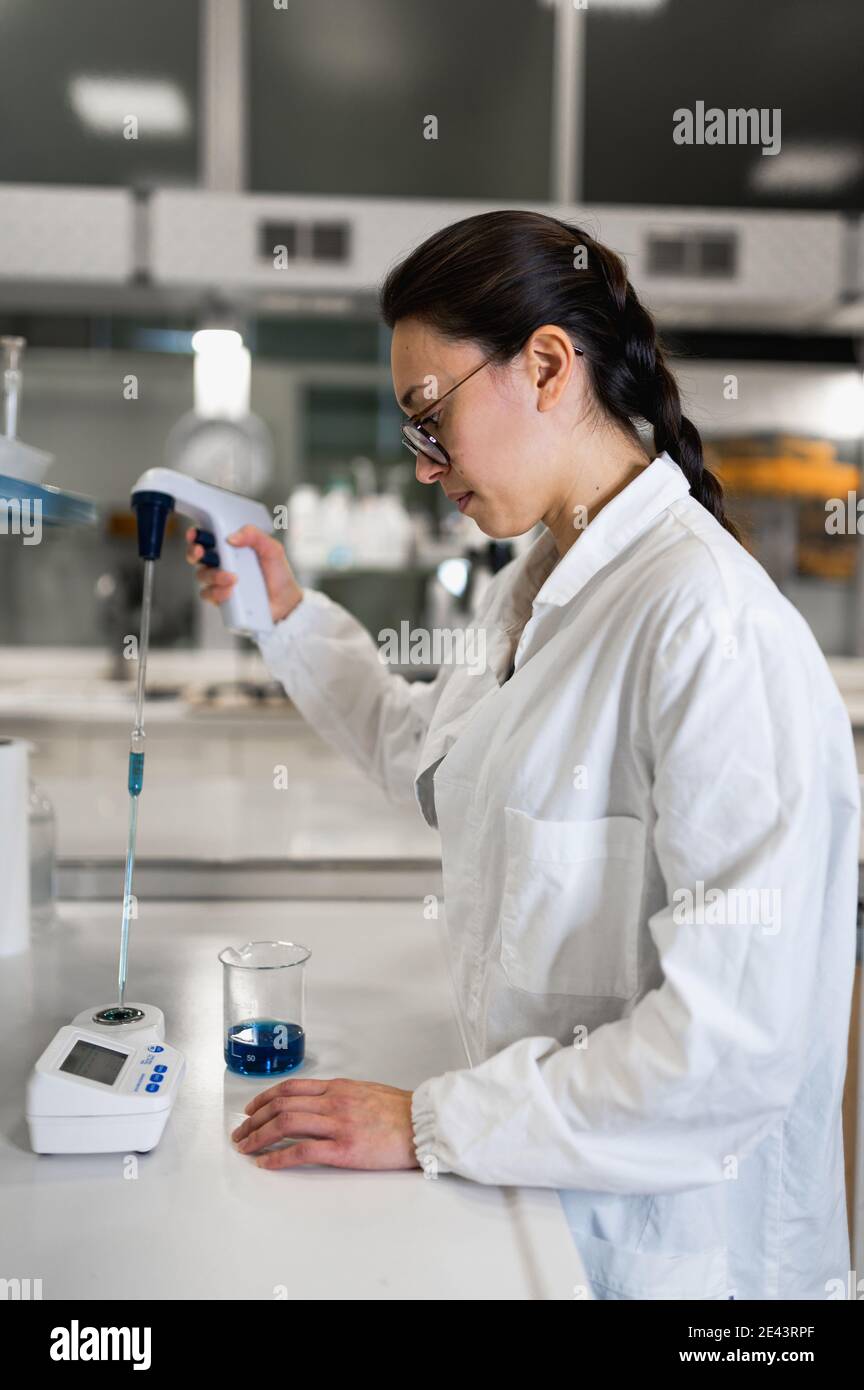 Side view of young female microbiologist in white coat dropping blue ...
