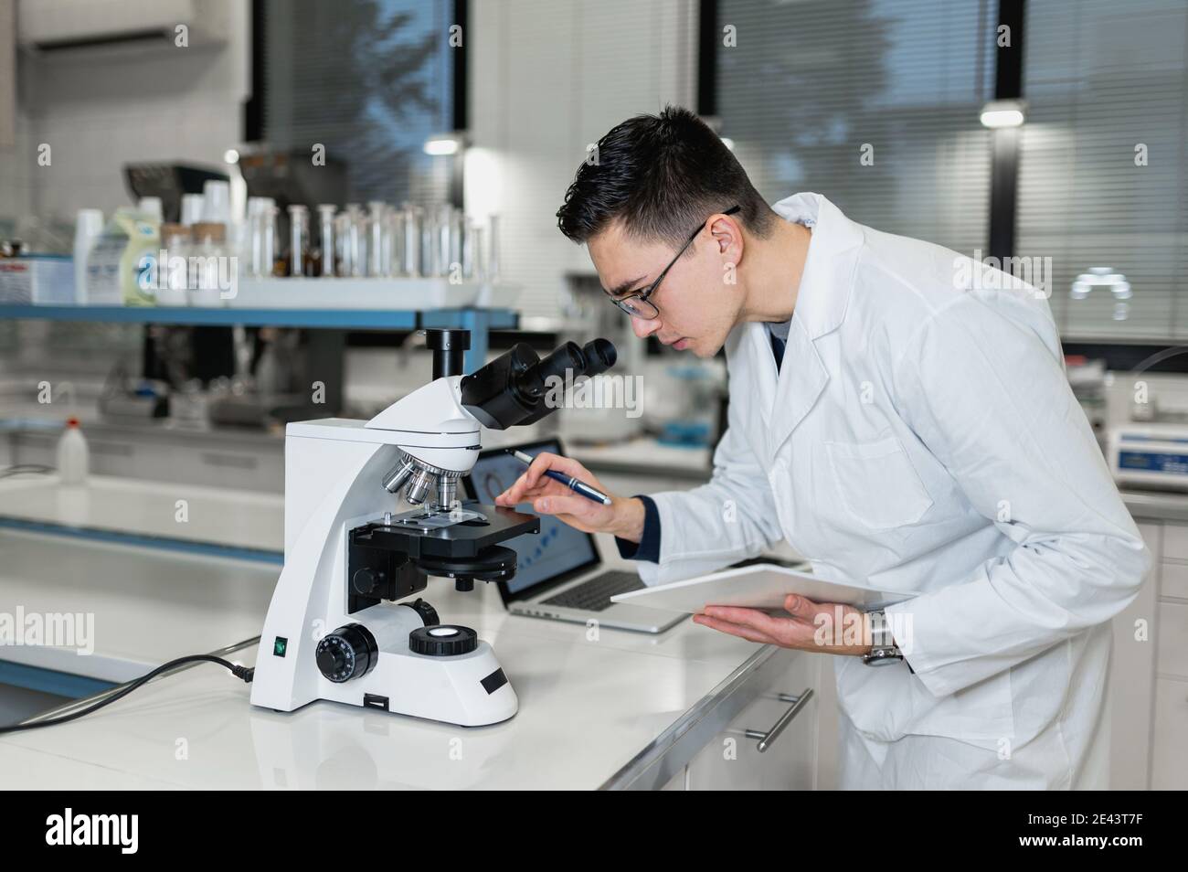 Side view of young male microbiologist in white coat examining chemical ...