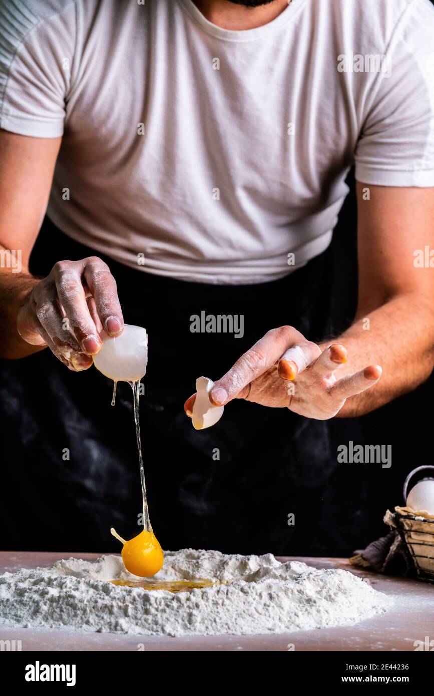 Faceless female baker breaking raw chicken egg into sifted flour pile ...