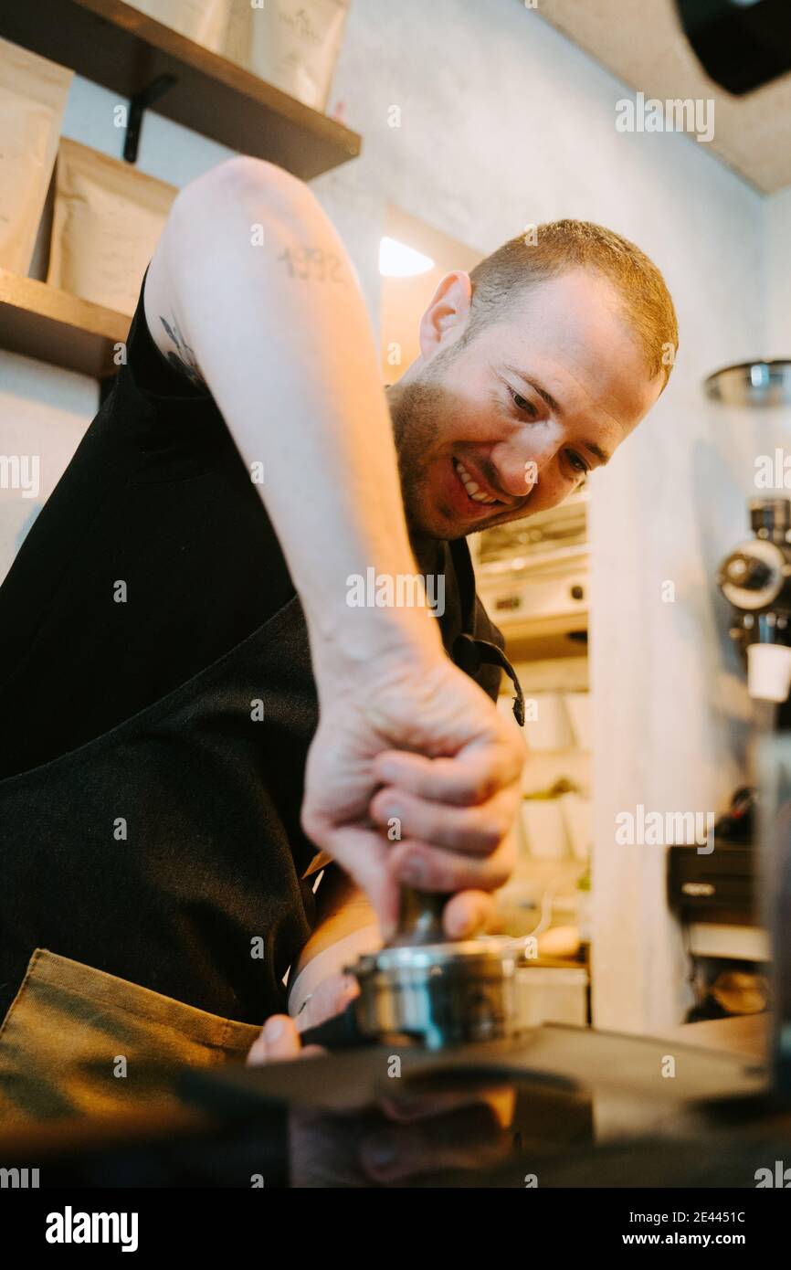 Low angle of smiling male barista pressing coffee in portafilter with ...