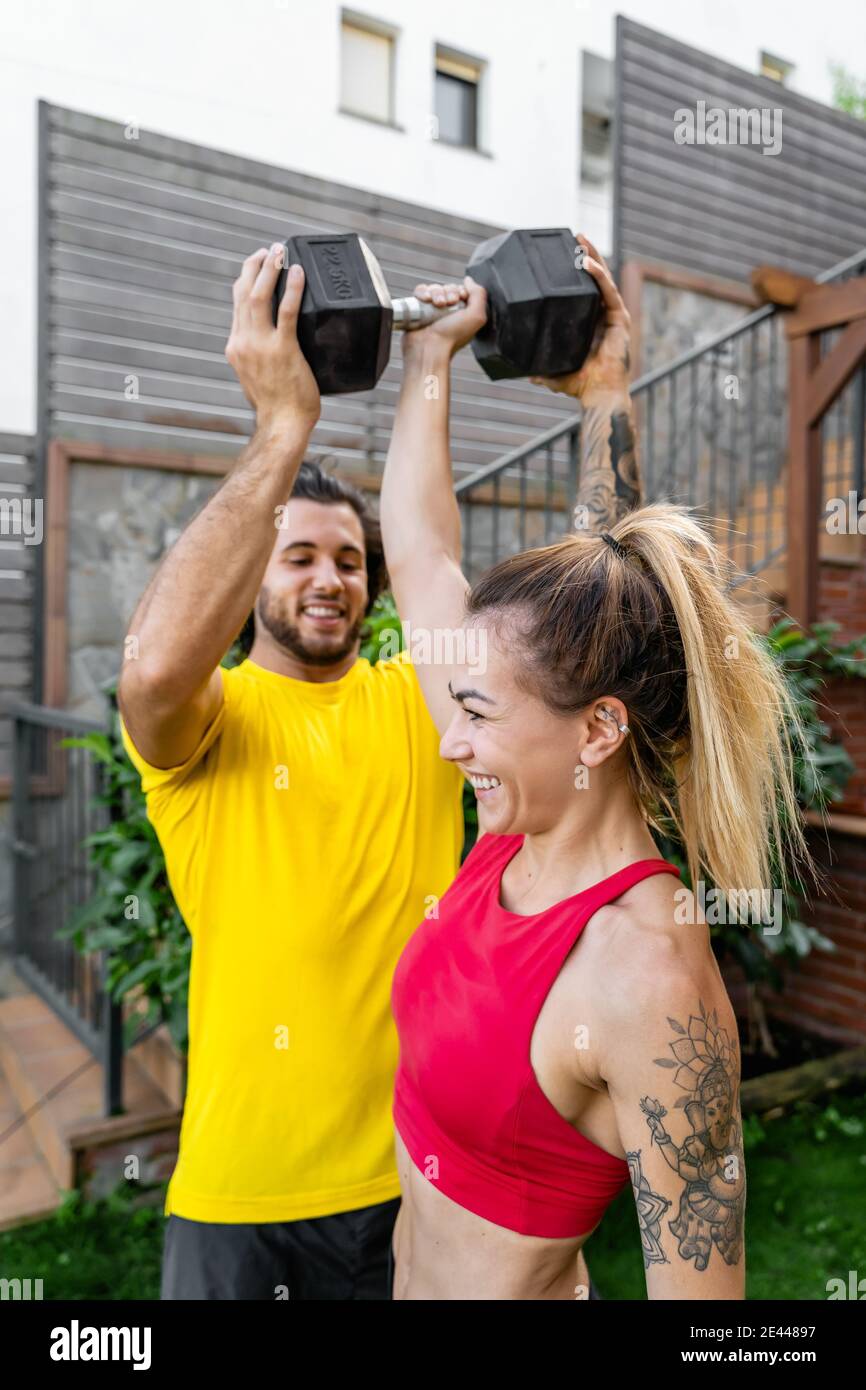 Cheerful athletic female lifting dumbbell with help of personal trainer ...