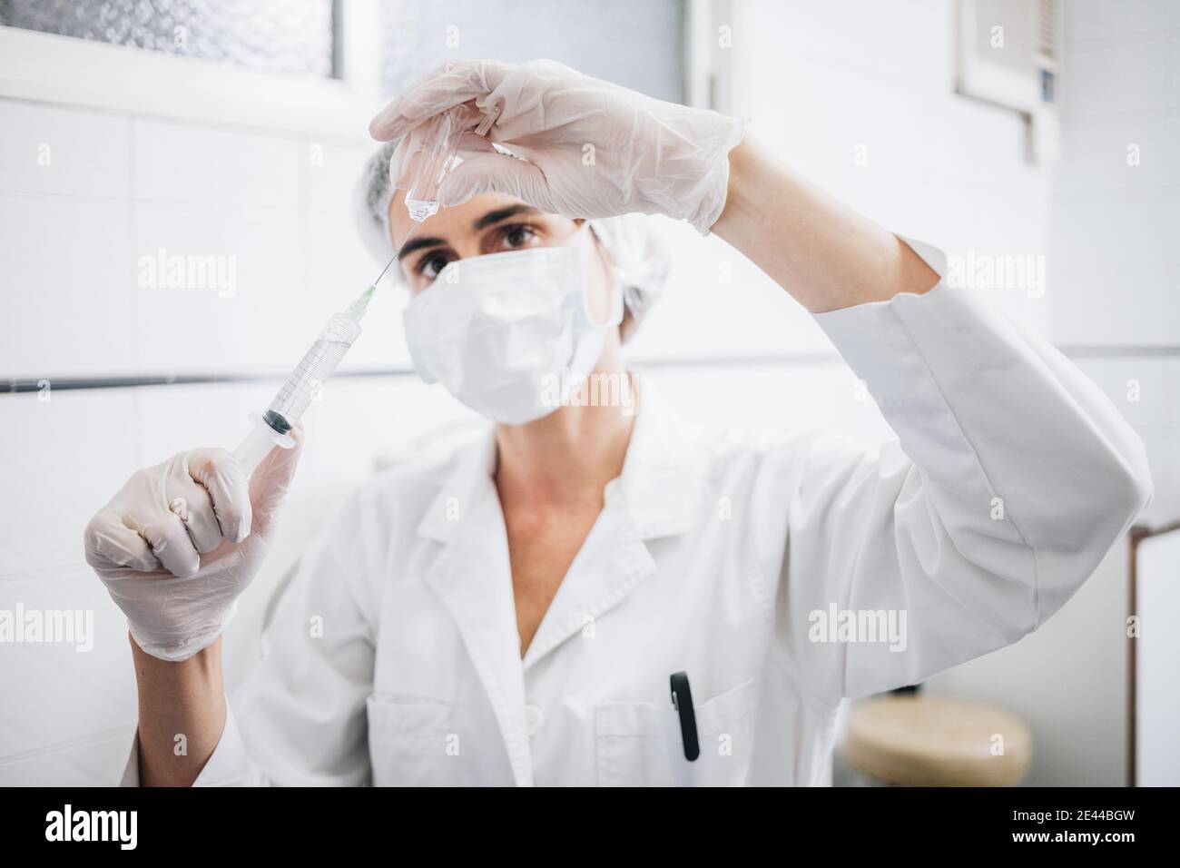 A nurse in uniform with mask, cap and gloves filling a needle with ...