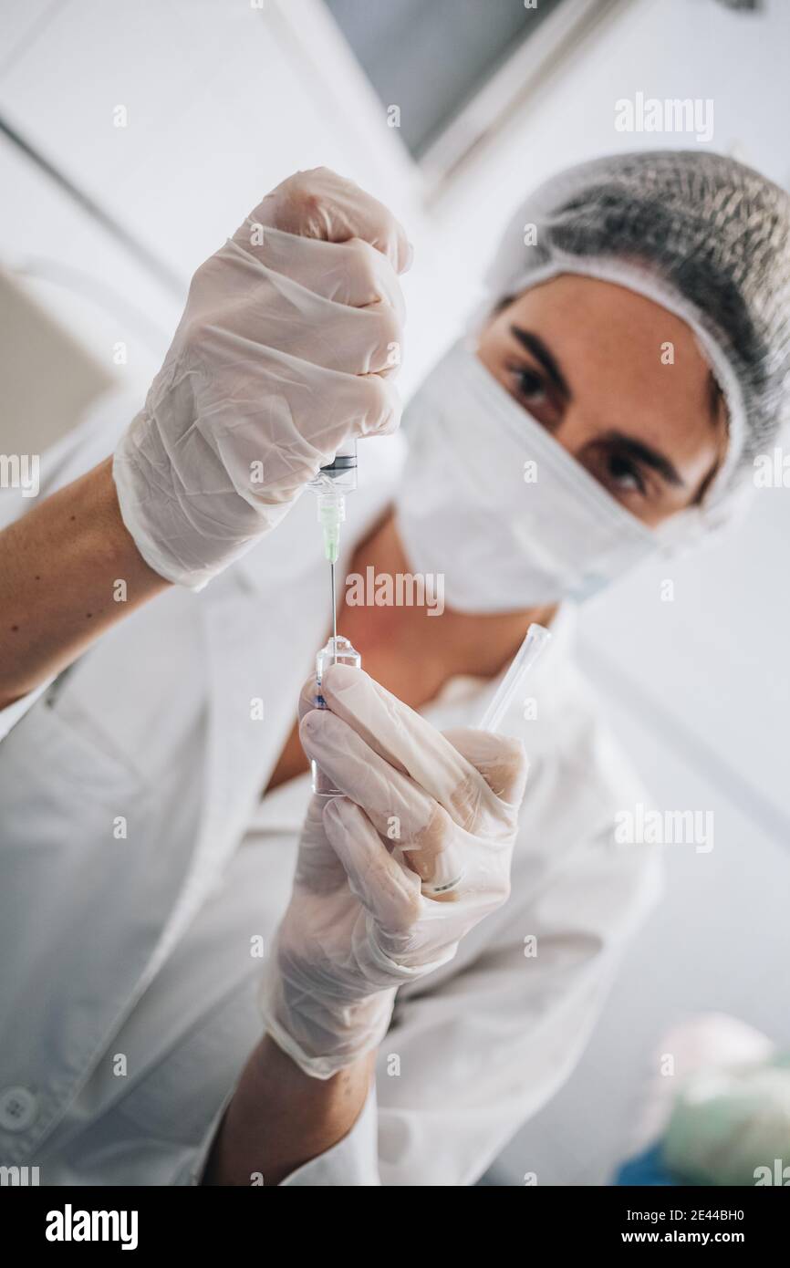 Vertical photo of a doctor with mask, cap and gloves filling a needle ...
