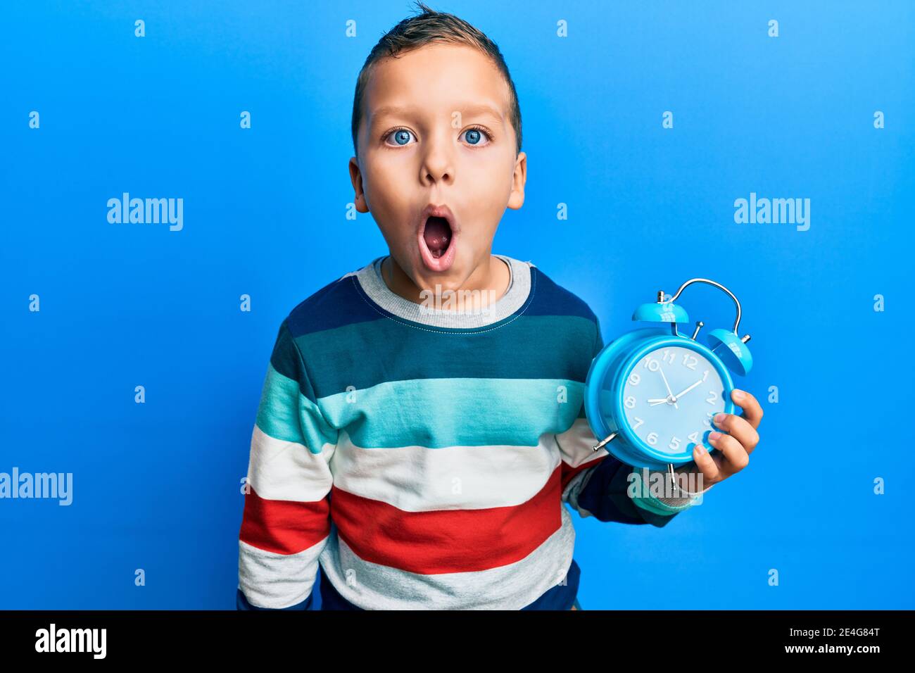 Little kid boy holding alarm clock scared and amazed with open mouth ...