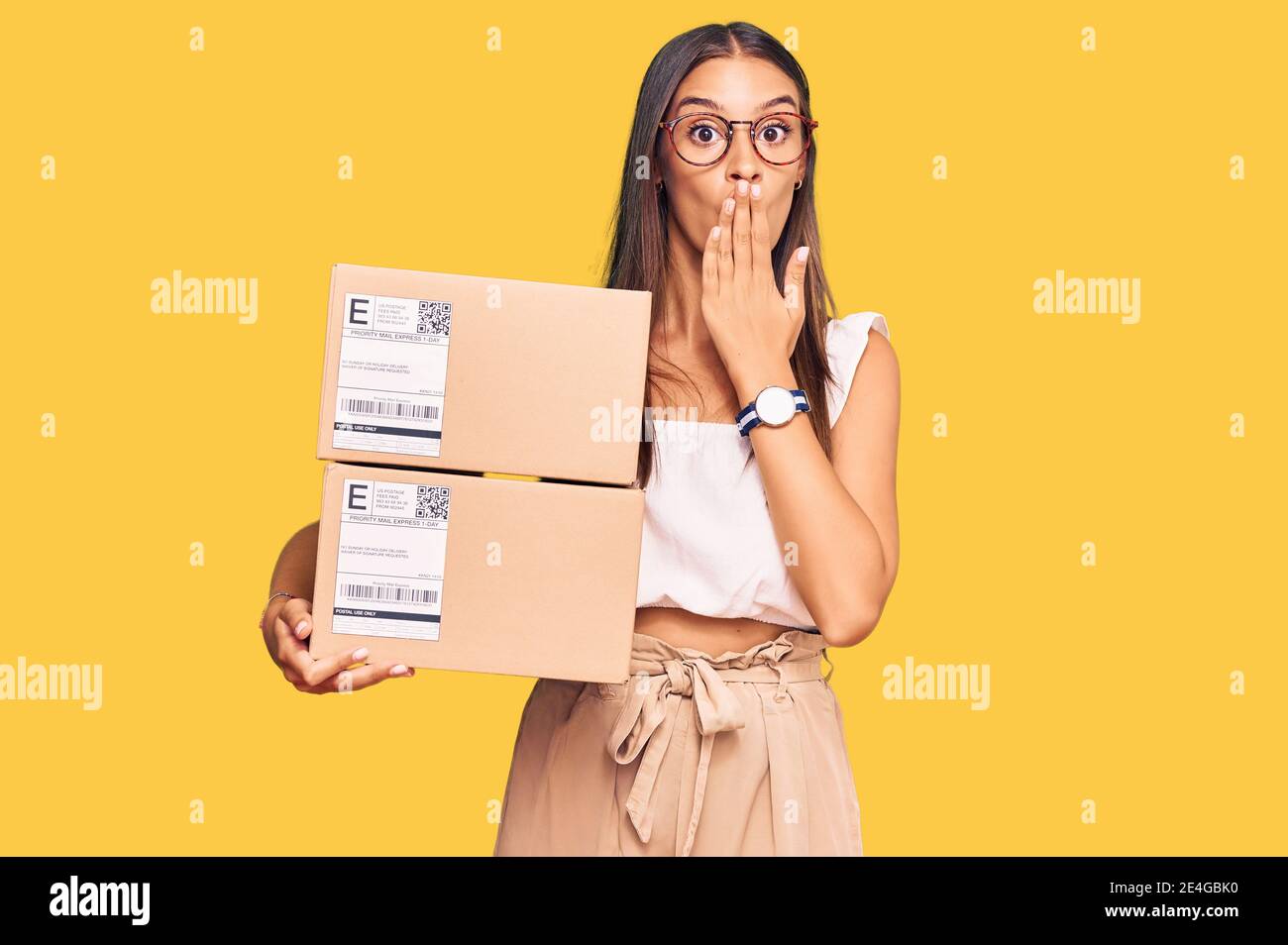 Young hispanic woman holding delivery package covering mouth with hand ...
