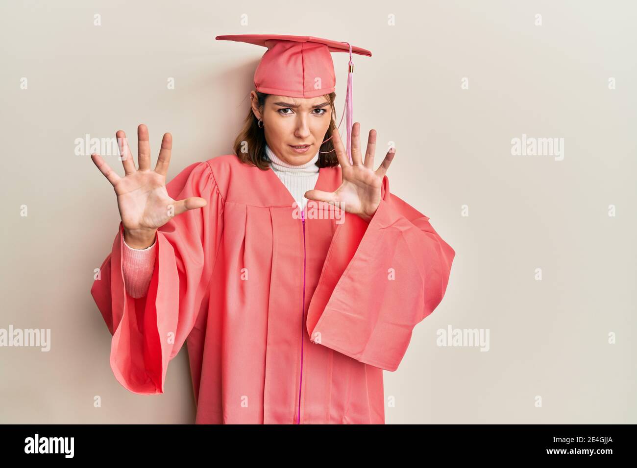 Young caucasian woman wearing graduation cap and ceremony robe afraid ...