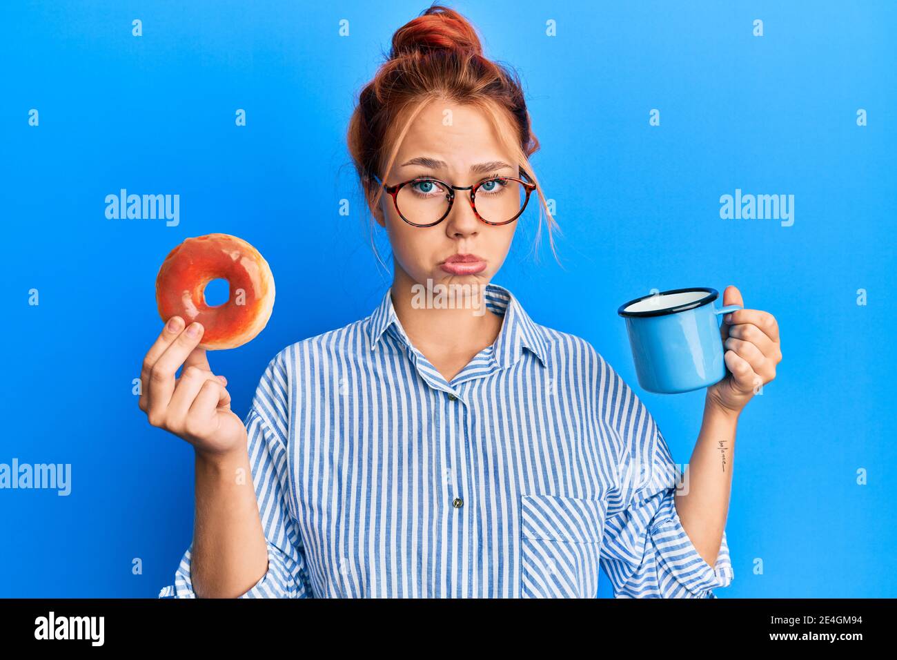 Young redhead woman eating breakfast holding chocolate donut and coffee ...