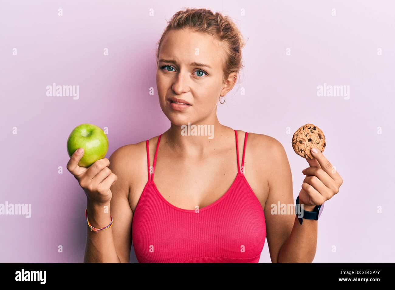 Beautiful caucasian woman holding green apple and cookie clueless and ...