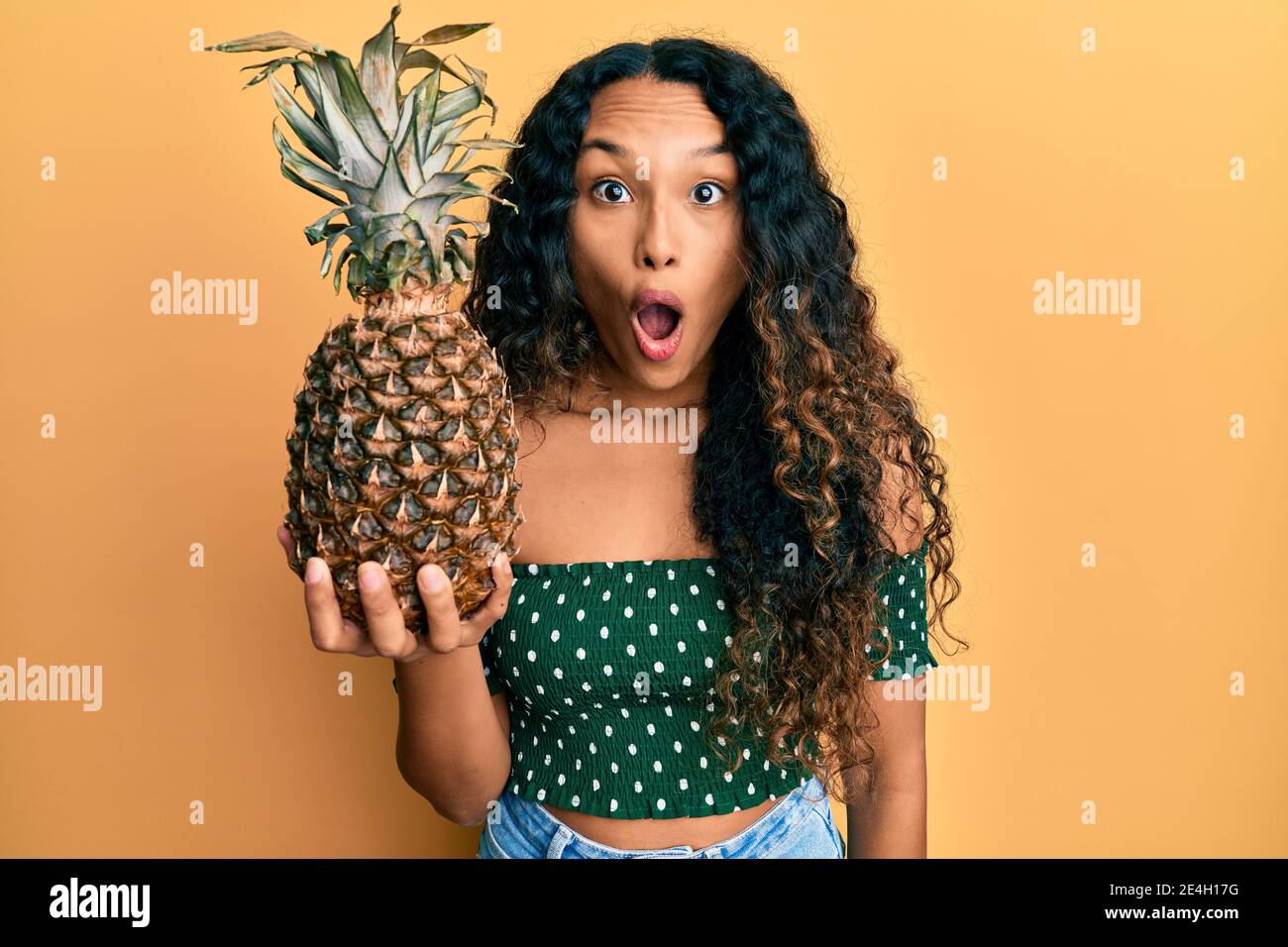 Young latin woman holding pineapple scared and amazed with open mouth ...