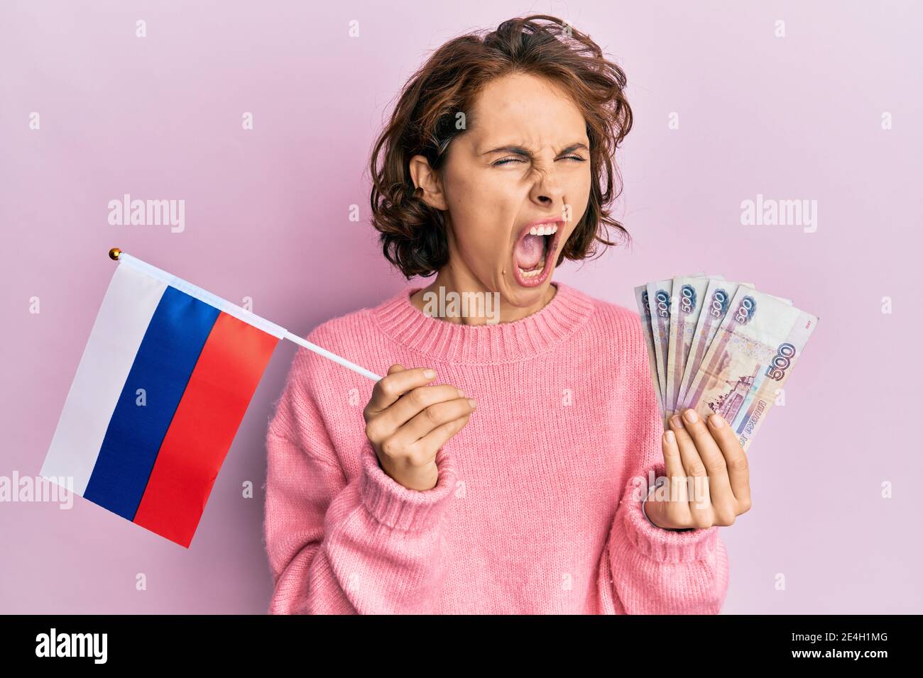 Young brunette woman holding russia flag and rubles banknotes angry and ...