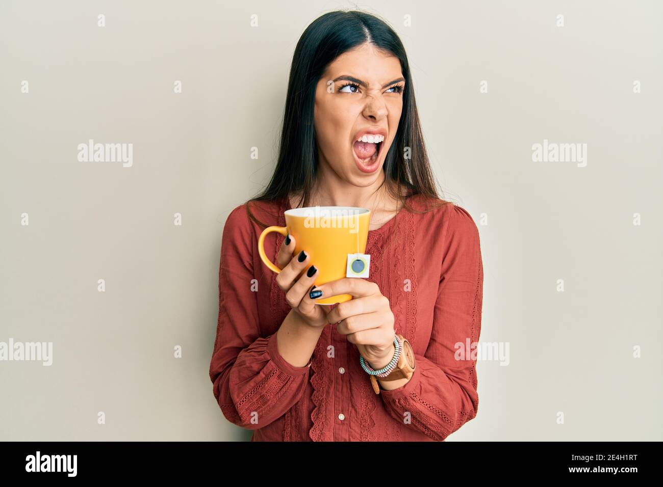 Young hispanic woman holding cup of tea angry and mad screaming ...