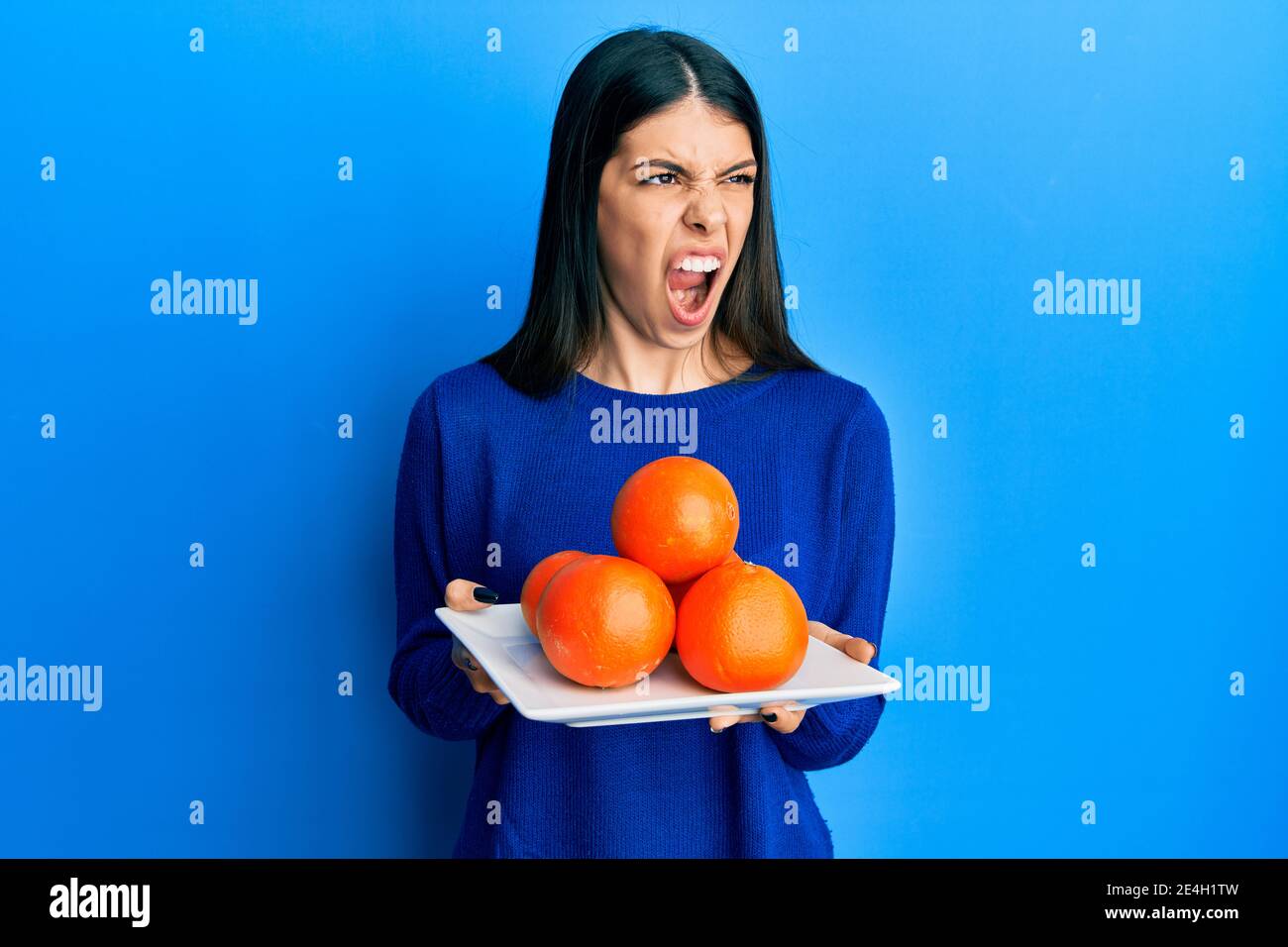 Young hispanic woman holding plate with fresh oranges angry and mad ...