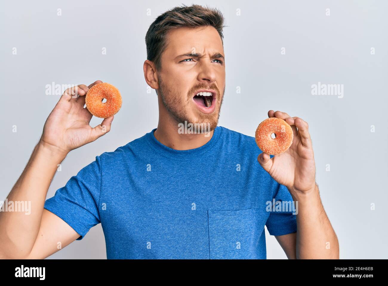 Handsome caucasian man eating donuts angry and mad screaming frustrated ...