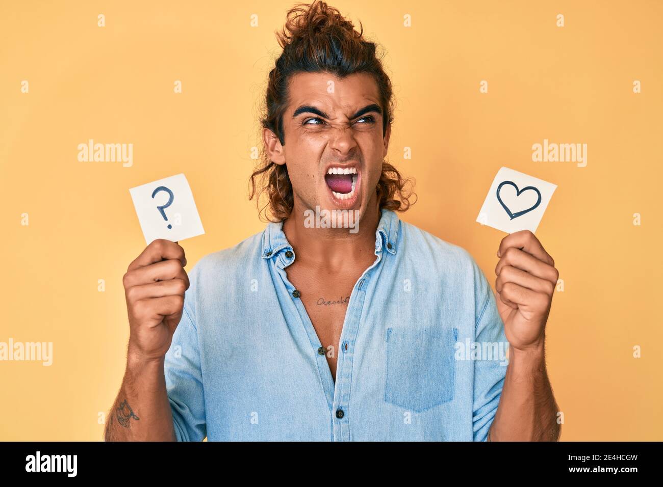 Young hispanic man holding heart and question mark reminder angry and ...