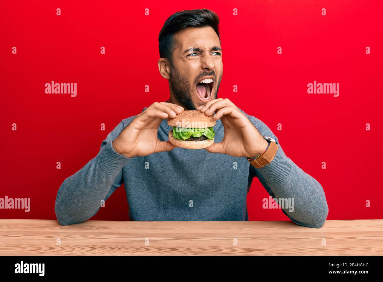 Handsome hispanic man eating a tasty classic burger angry and mad ...
