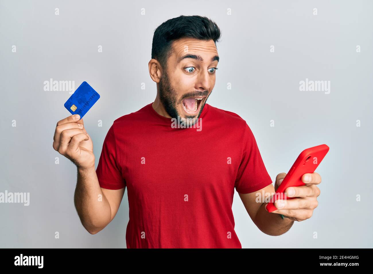 Young hispanic man holding smartphone and credit card celebrating crazy ...
