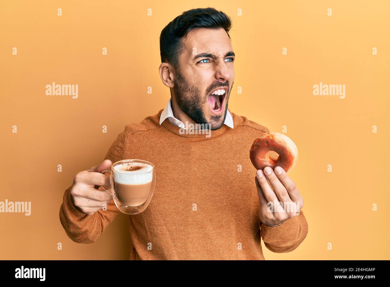 Young hispanic man eating doughnut and drinking coffee angry and mad ...