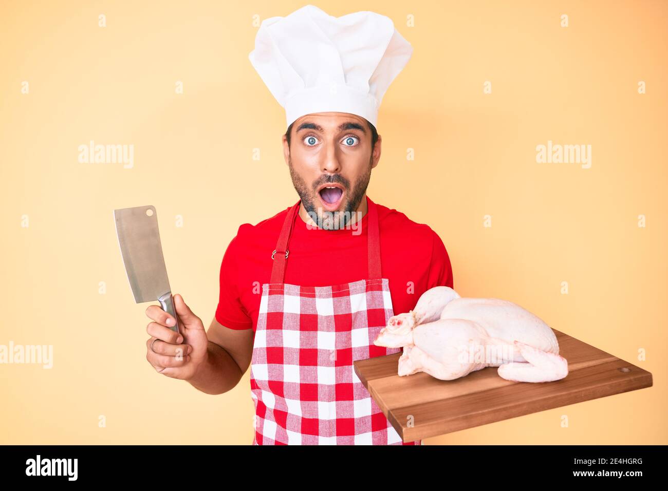 Young hispanic man cooking chicken holding knife afraid and shocked ...