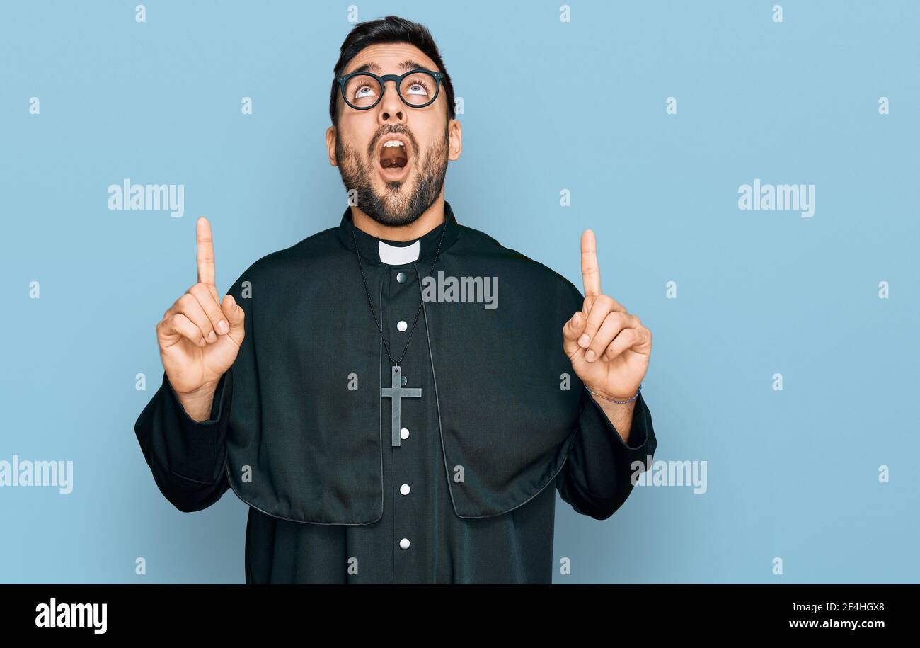 Young hispanic man wearing priest uniform amazed and surprised looking ...